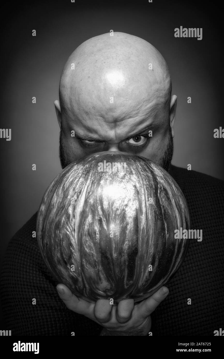 A bald man holds a bowling ball in front of his face squinting at the ...