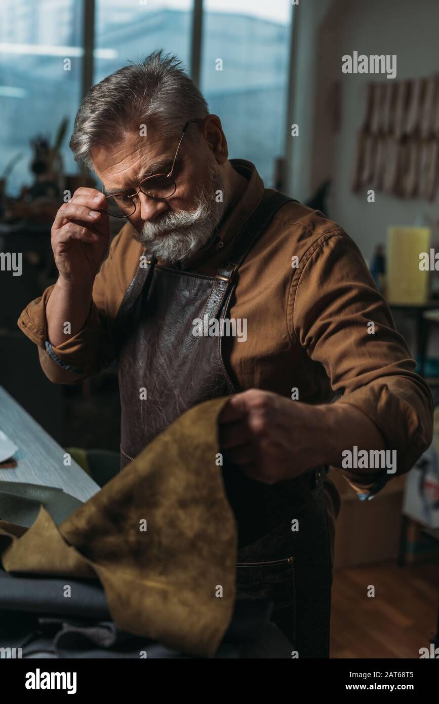 attentive shoemaker touching glasses while looking at piece of genuine ...