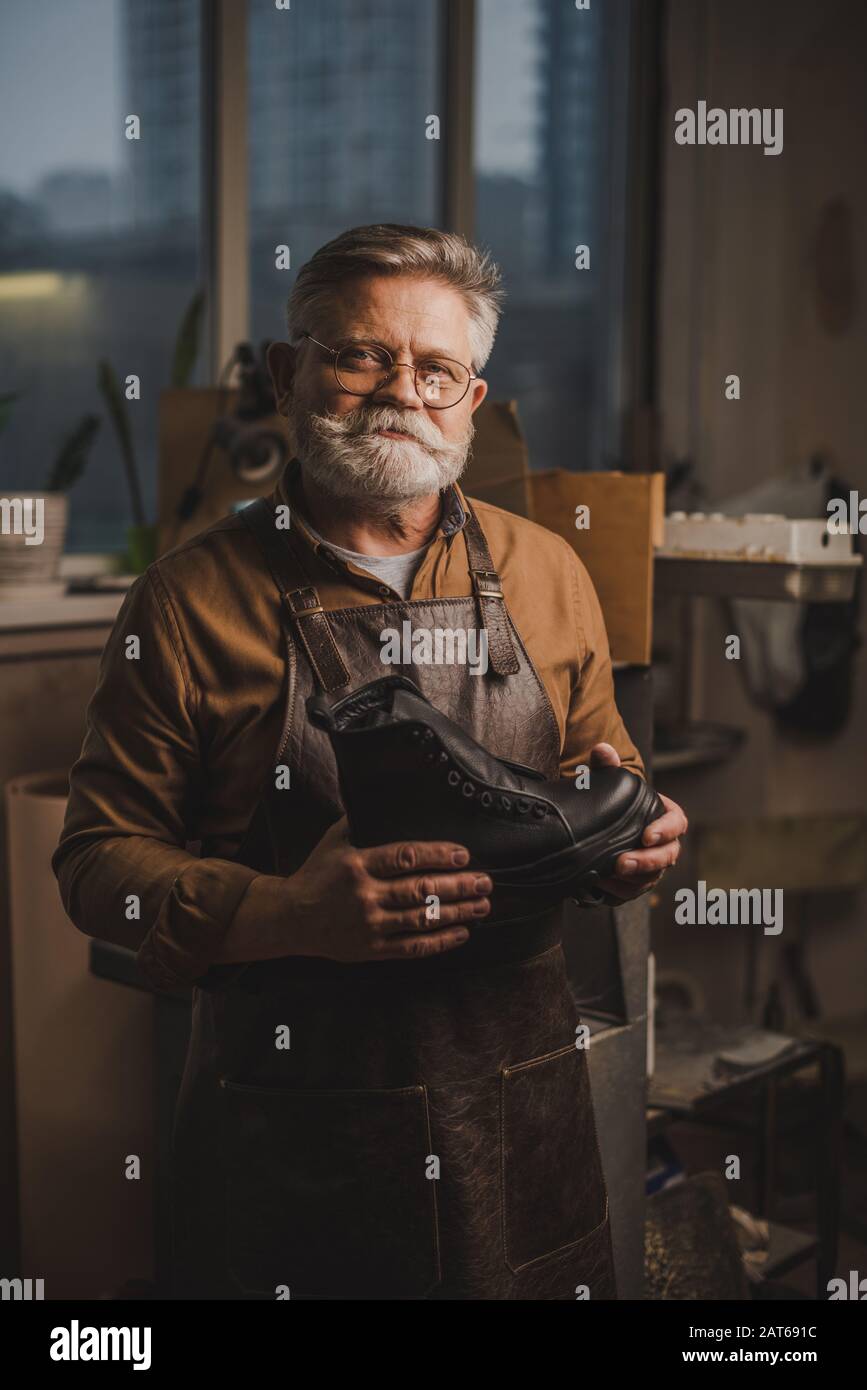 positive, senior shoemaker smiling at camera while holding leather boot ...