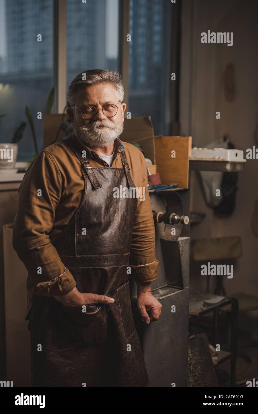 positive, bearded shoemaker in leather apron looking at camera while ...