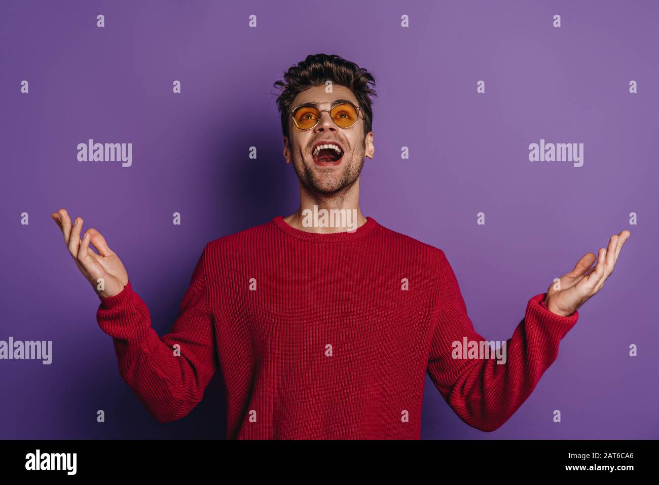 happy man looking up while standing with open arms on purple background ...