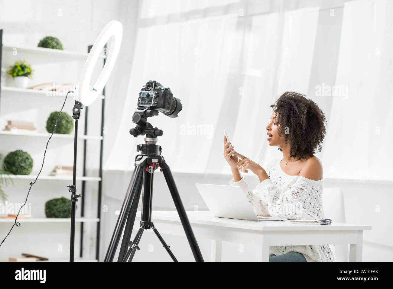 attractive african american influencer holding smartphone near laptop ...