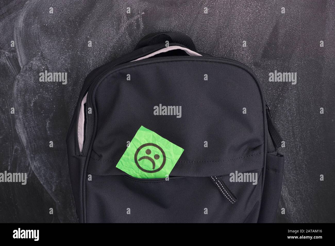 Backpack with a sad face on paper note standing against blackboard ...