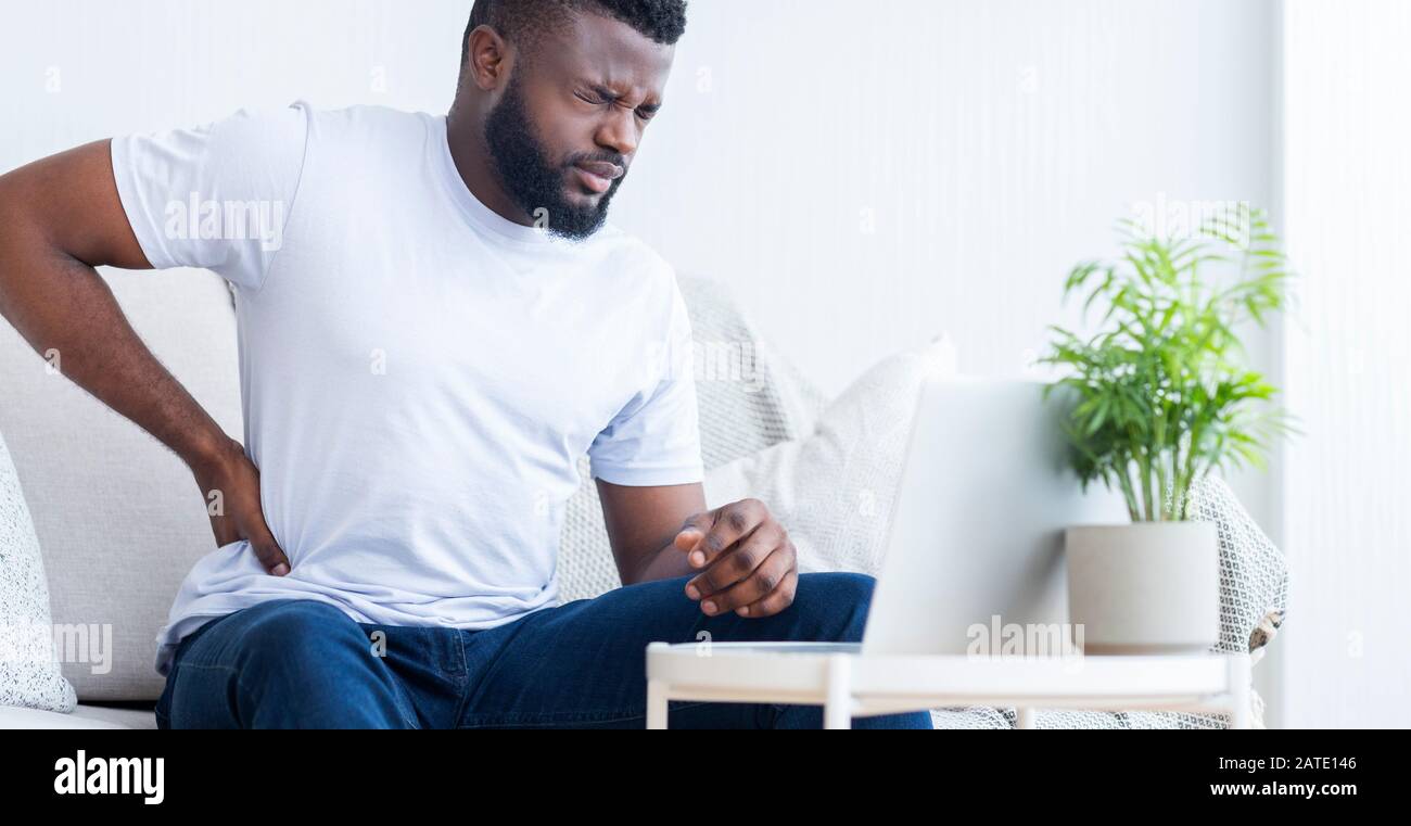 Young black man touching his sore back, working on laptop Stock Photo ...