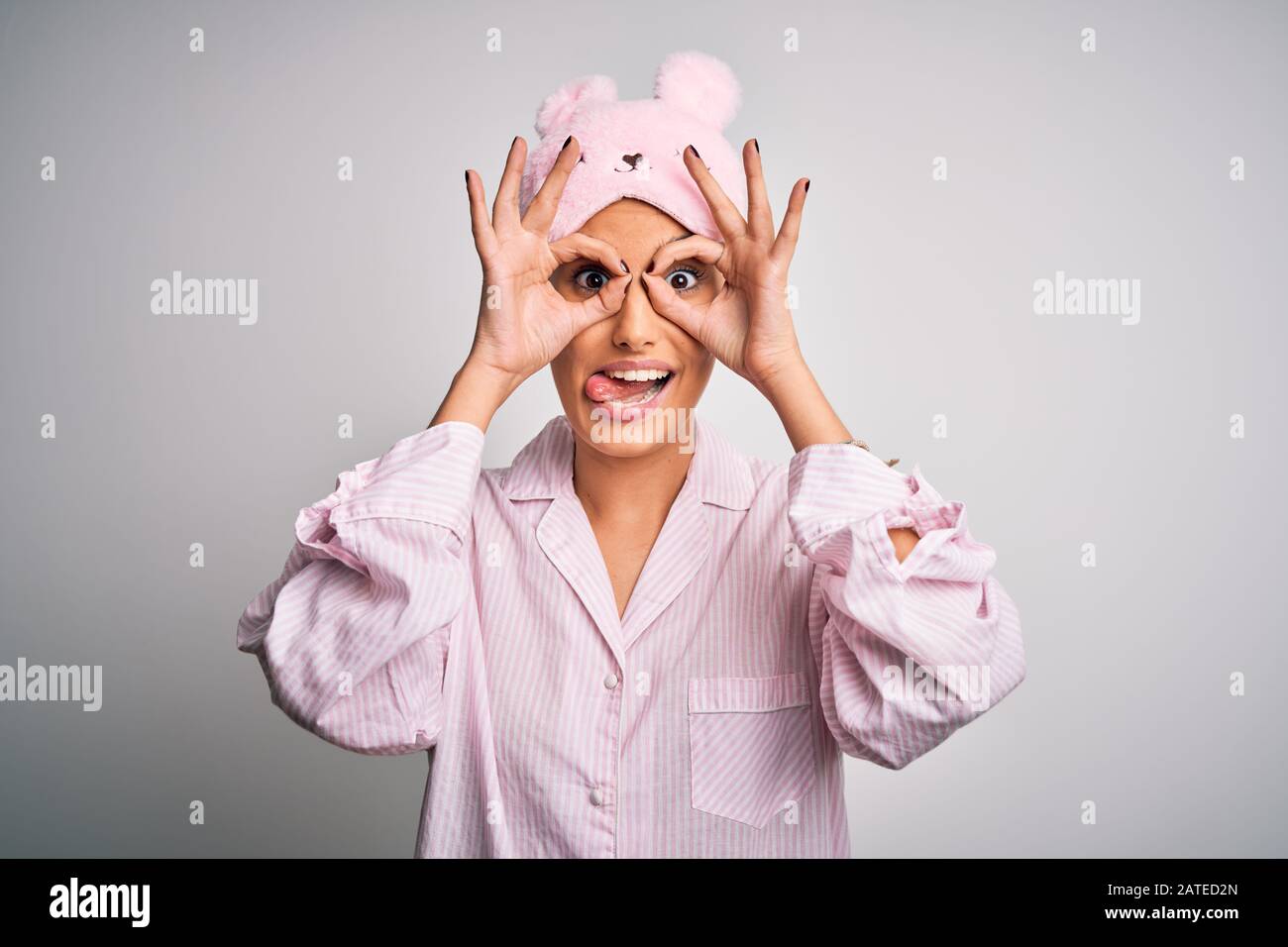 Young beautiful brunette woman wearing pajama and sleep mask over white background doing ok ...