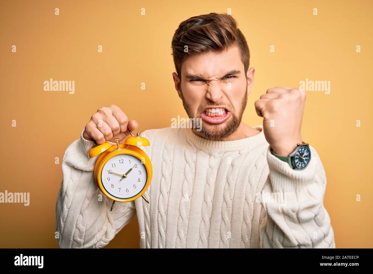 Young blond man with beard and blue eyes holding alarm clock over ...