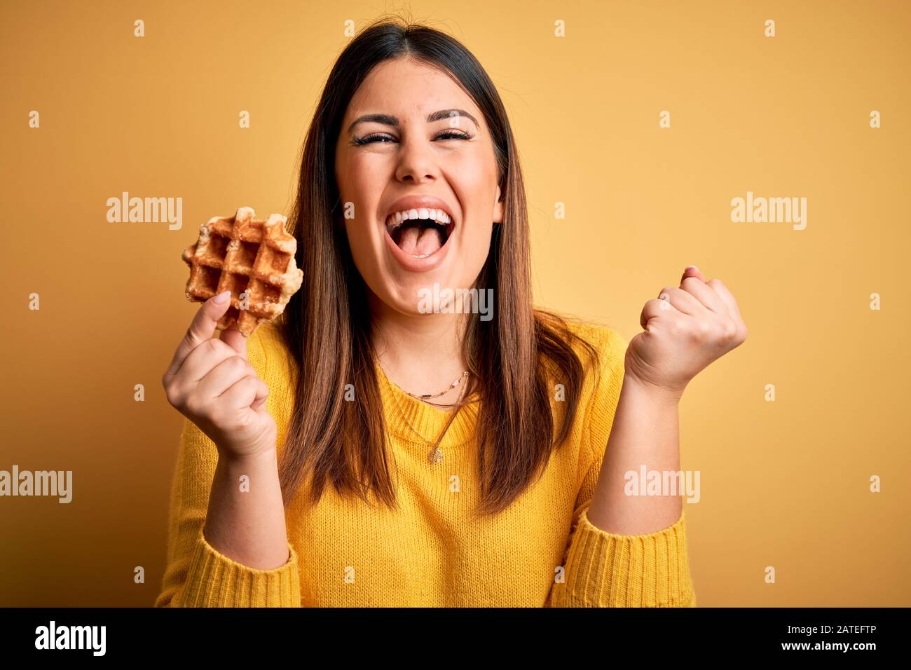 Young beautiful woman eating sweet waffle pastry over yellow background ...