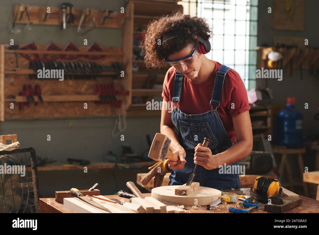 Concentrated curly woman in ear defenders and goggles using hammer and ...
