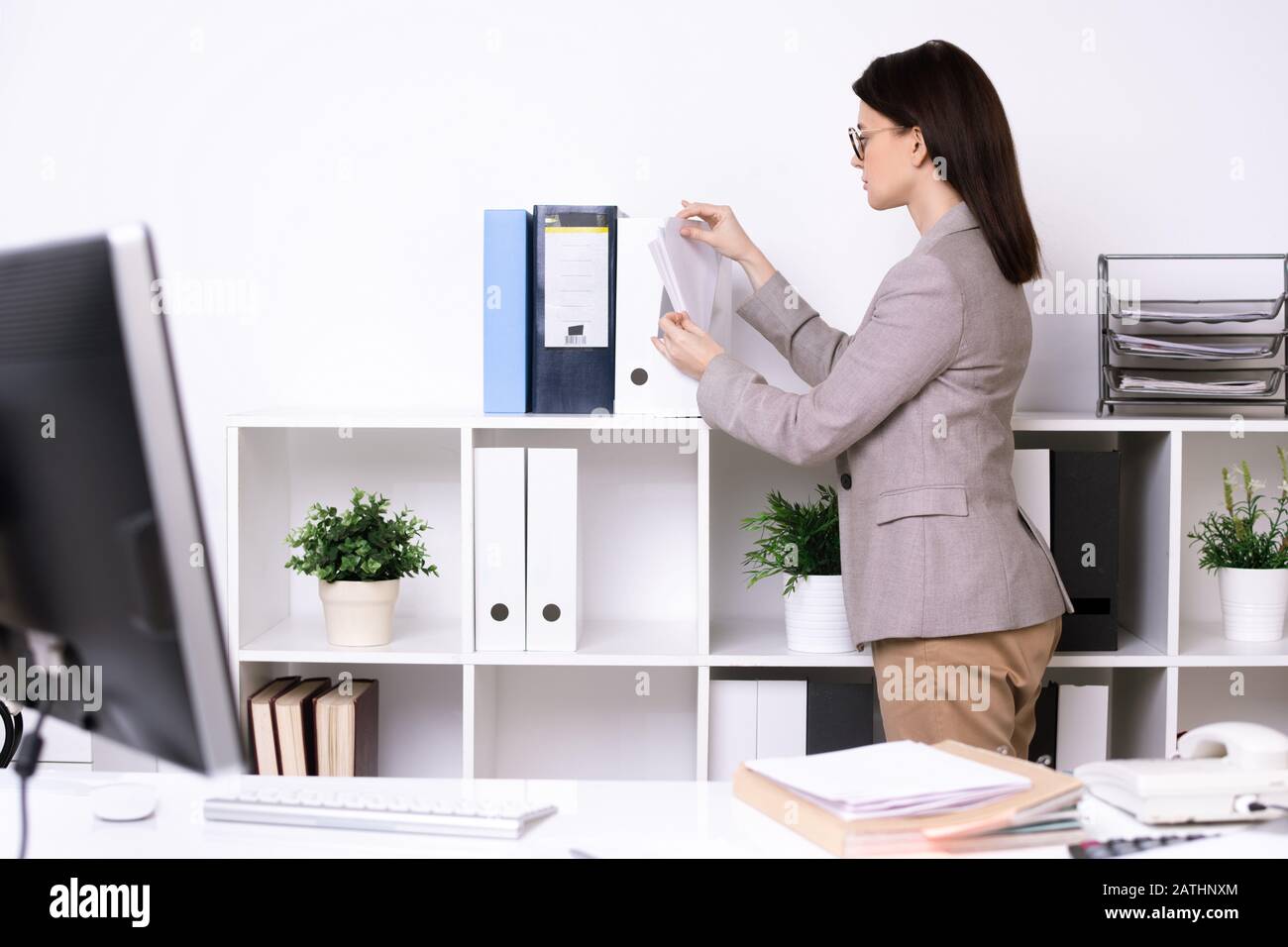Brunette secretary in jacket cleaning up office room and sorting files ...