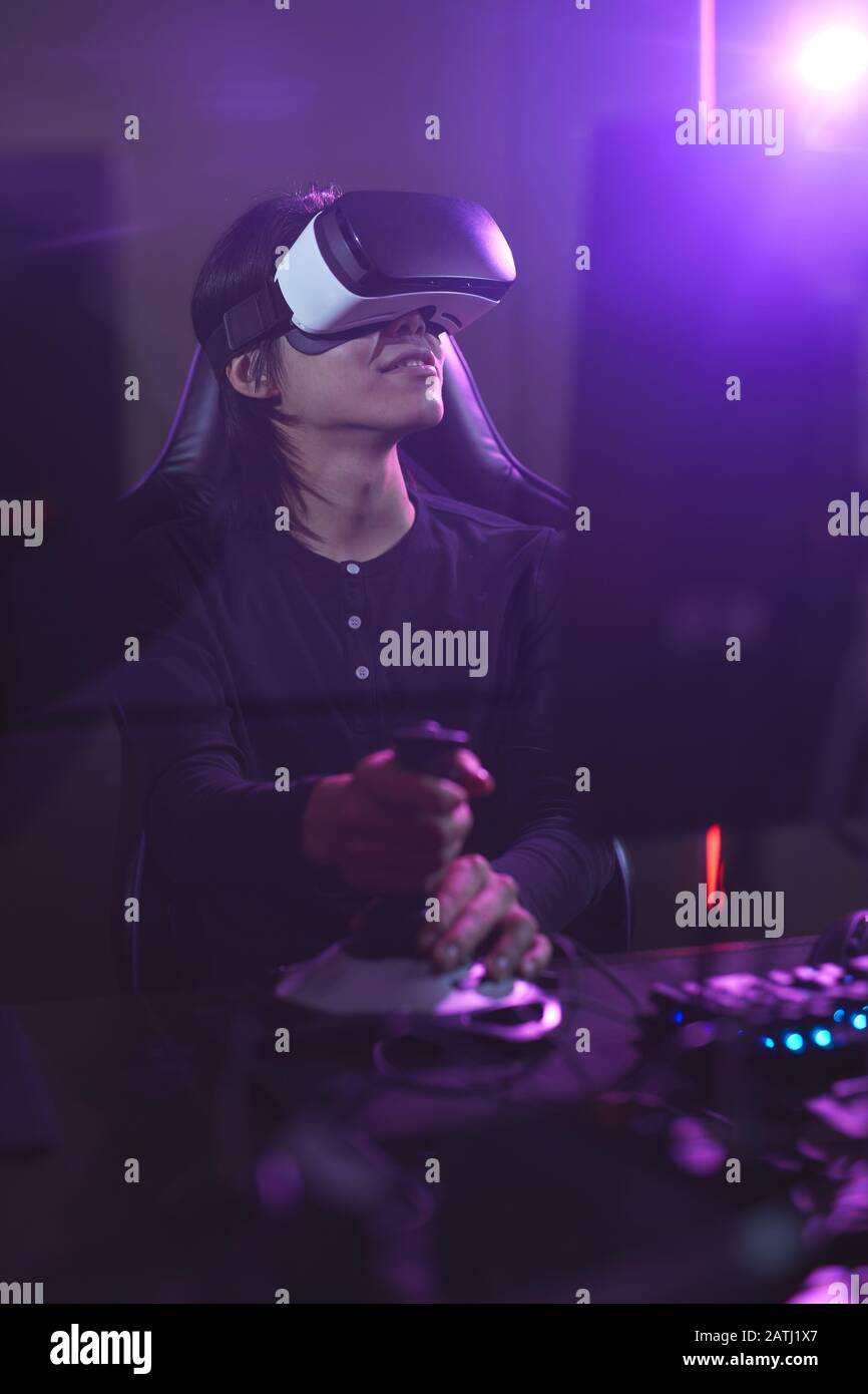 Vertical portrait of young Asian man wearing VR headset while playing videogames using racing ...