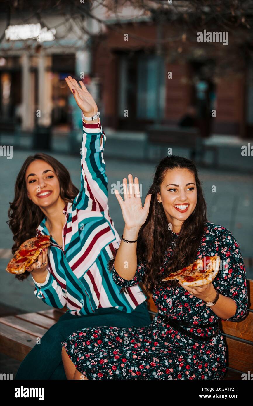 Two beautiful young women eating pizza outdoors and waving their ...