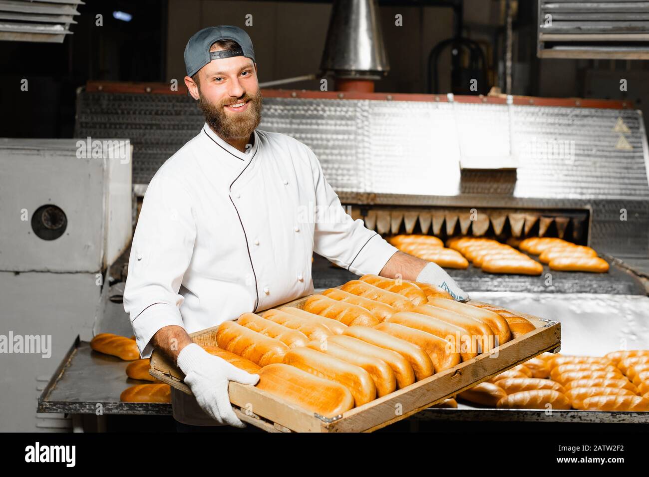 A baker holds a tray with fresh hot bread against the background of an industrial oven in a ...