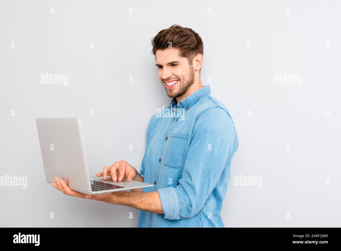 Smiling young man holding laptop and typing while standing against grey ...