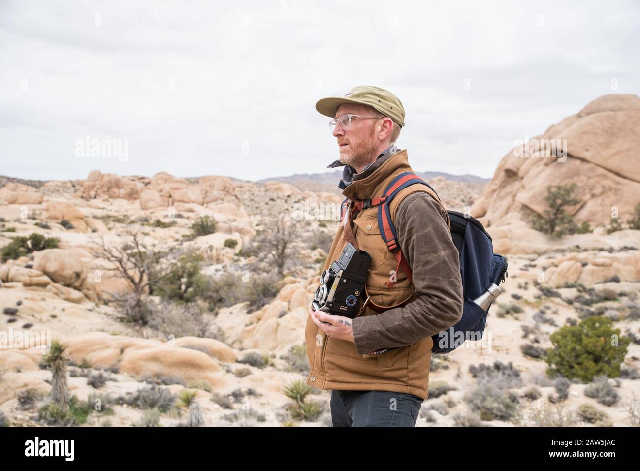Man with glasses, hat, and camera in desert by stones and plant life ...