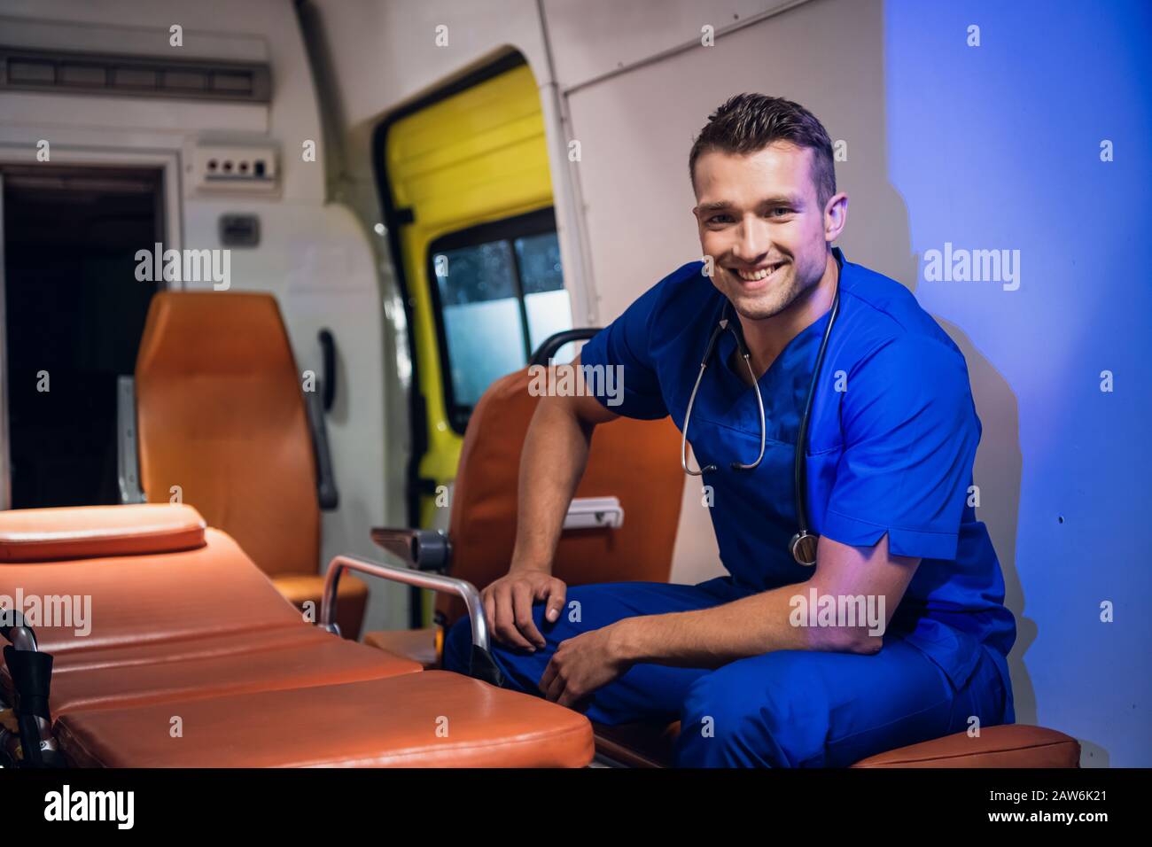 Young man in blue medical uniform sits in the ambulance car and smiles ...