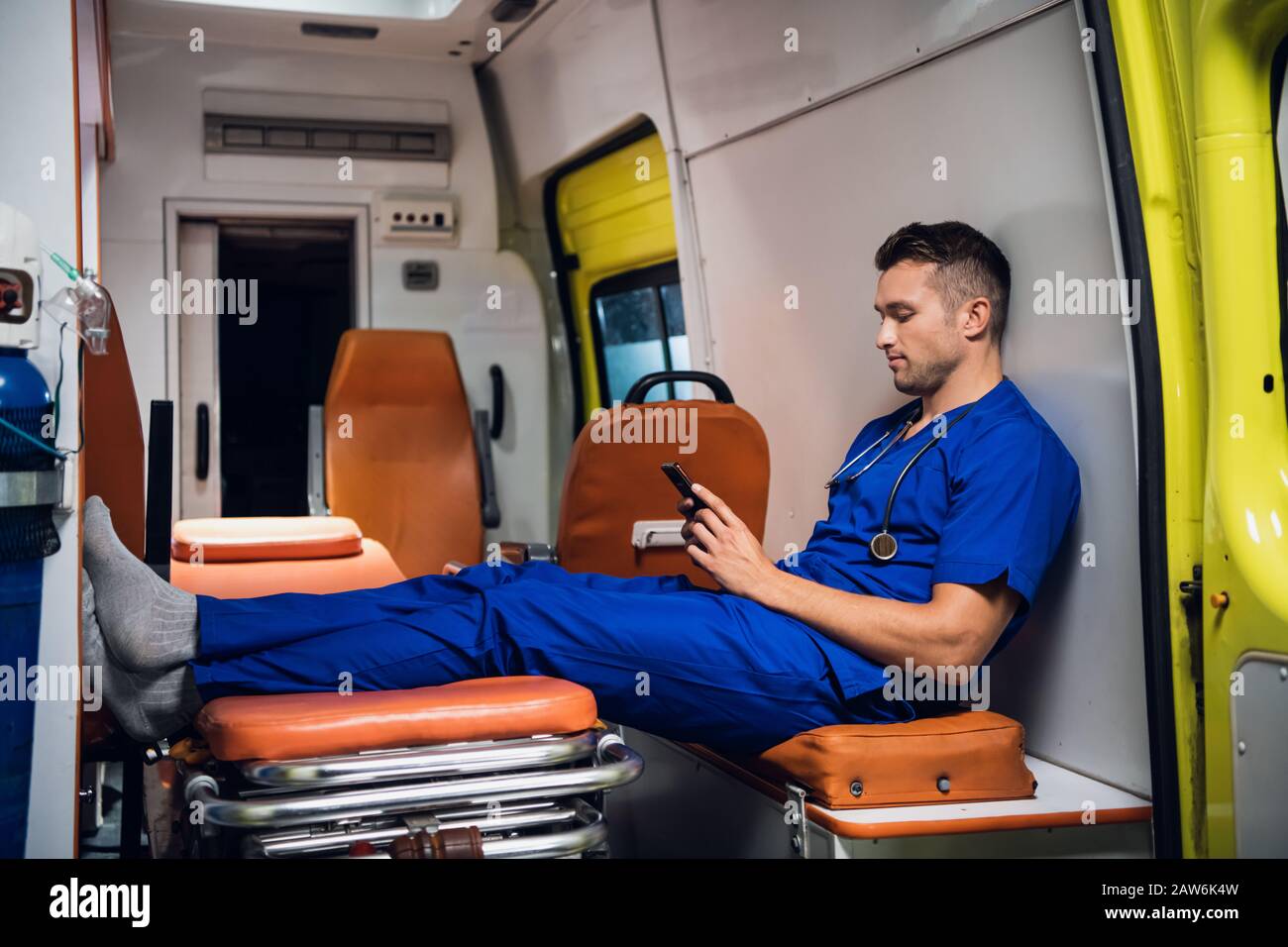 Young corpsman in blue medical uniform sits with phone in the ambulance ...