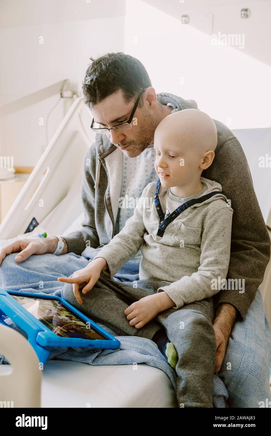 Father recovering from surgery watches tablet with son on hospital bed ...