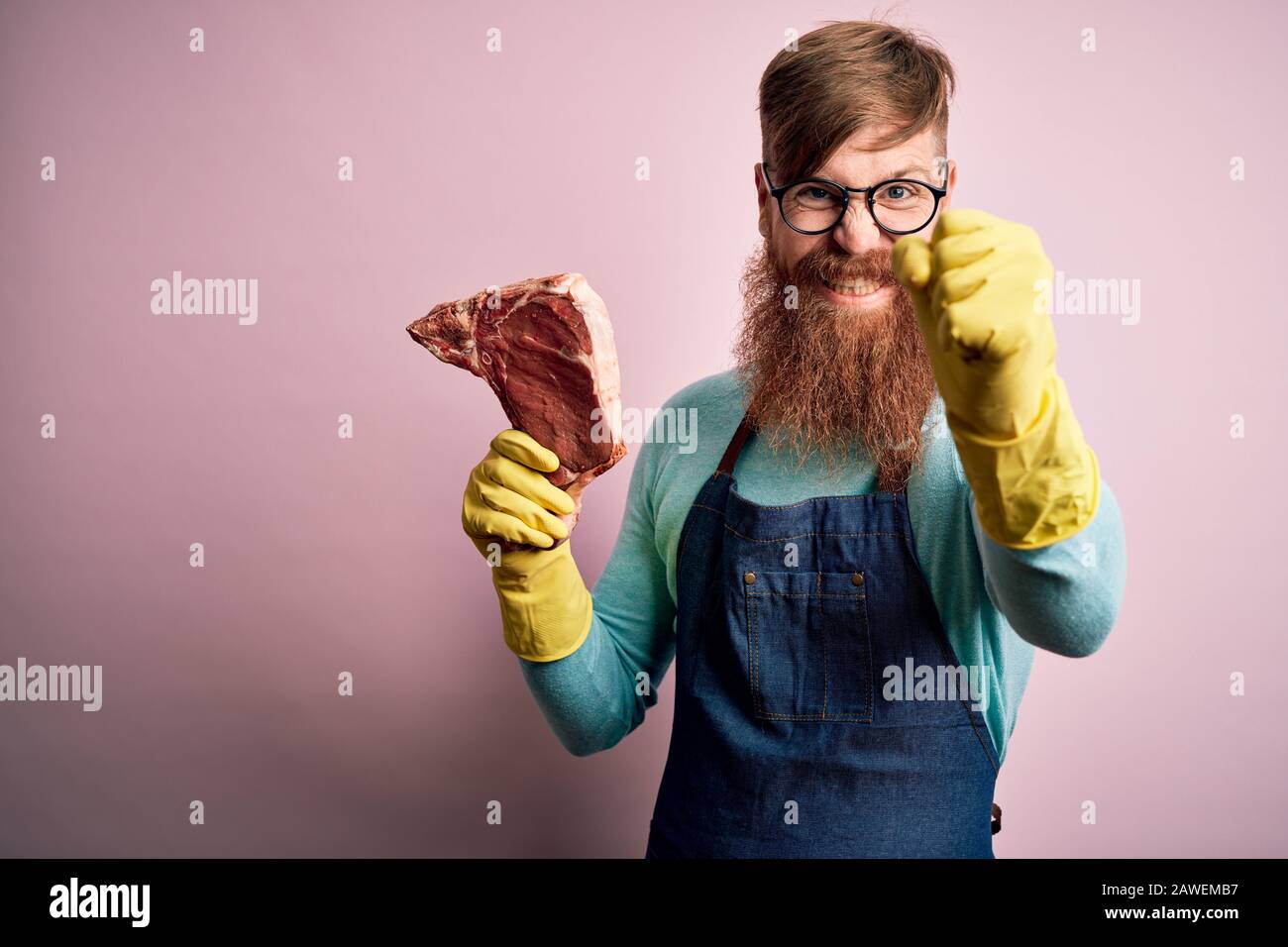 Redhead Irish butcher man with beard holding raw beef steak over pink ...