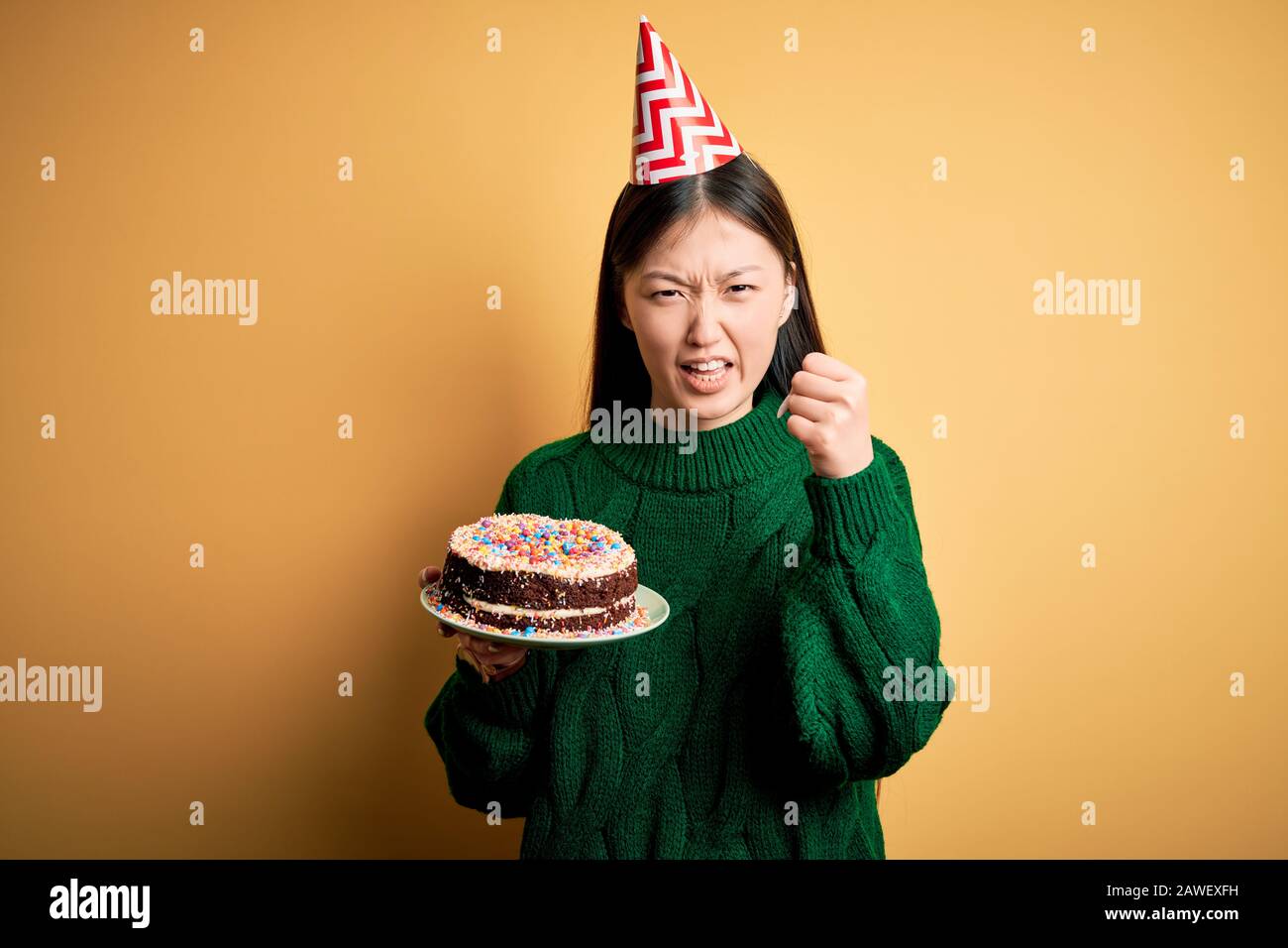Young asian woman holding birthday cake wearing party hat over yellow ...