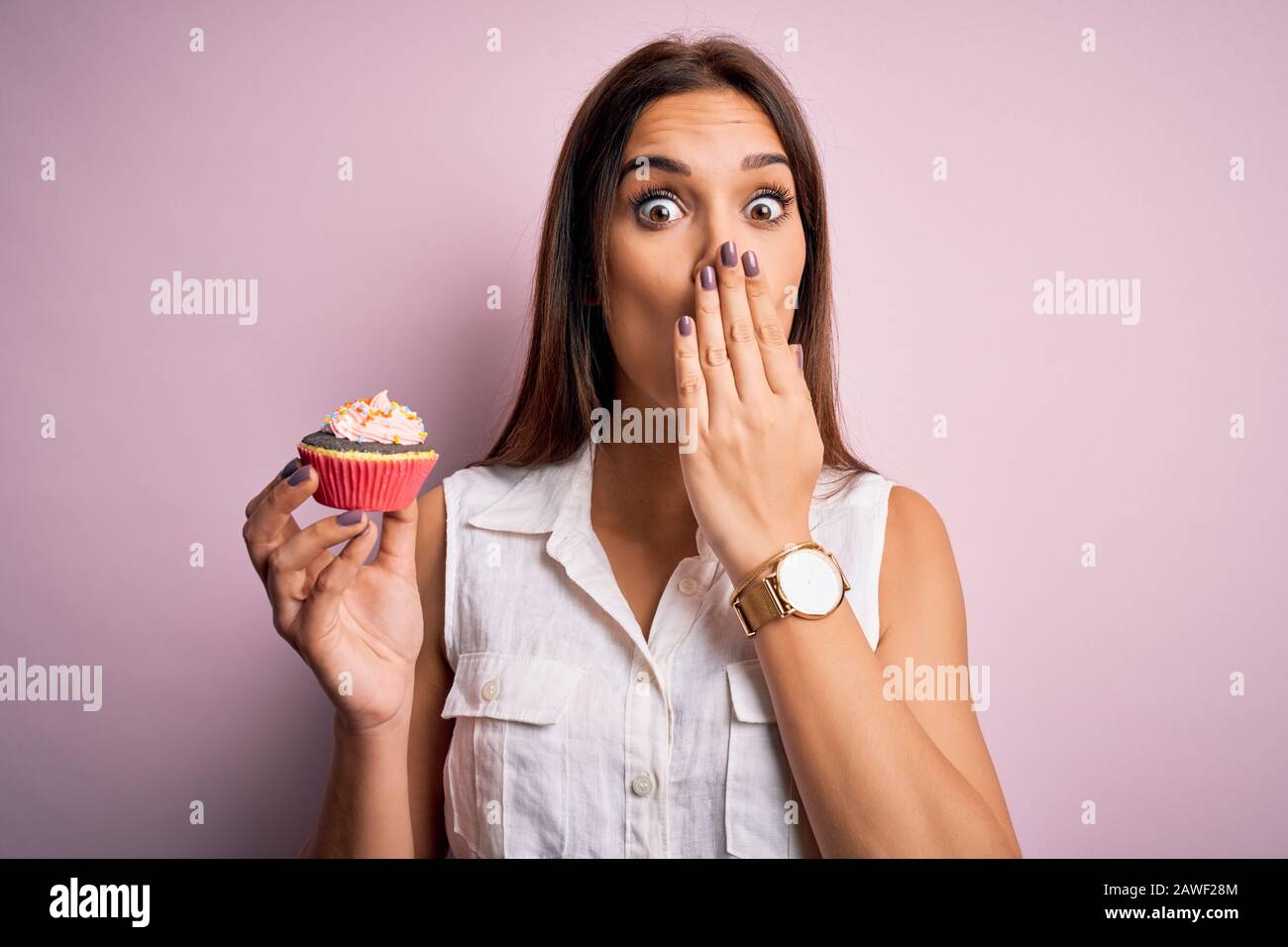 Young beautiful brunette woman eating chocolate cupcake over isolated pink background cover ...
