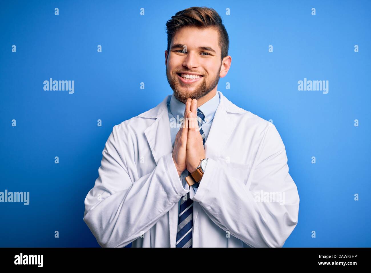 Young blond therapist man with beard and blue eyes wearing coat and tie over background praying ...