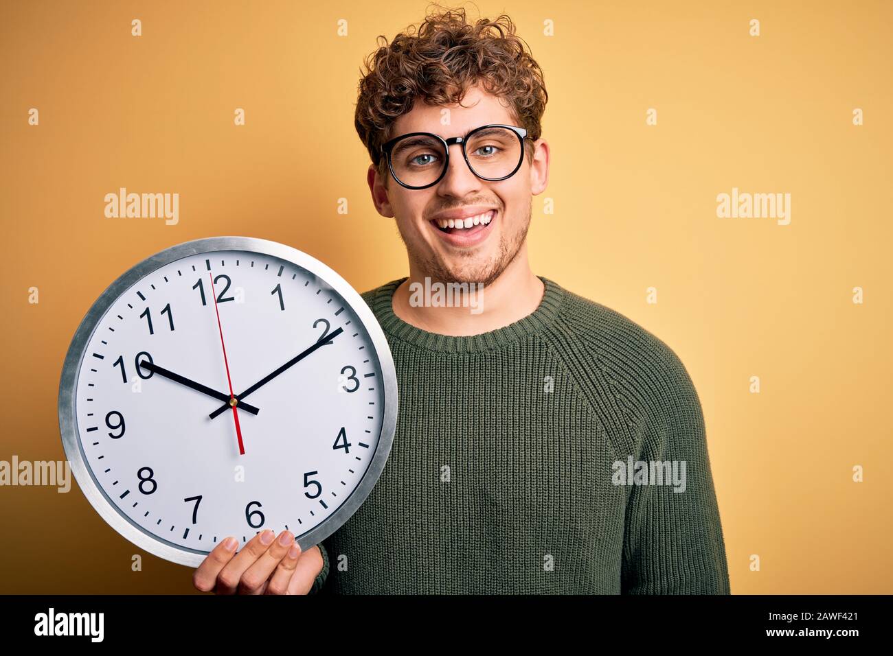 Young blond man with curly hair wearing glasses holding big clock over ...