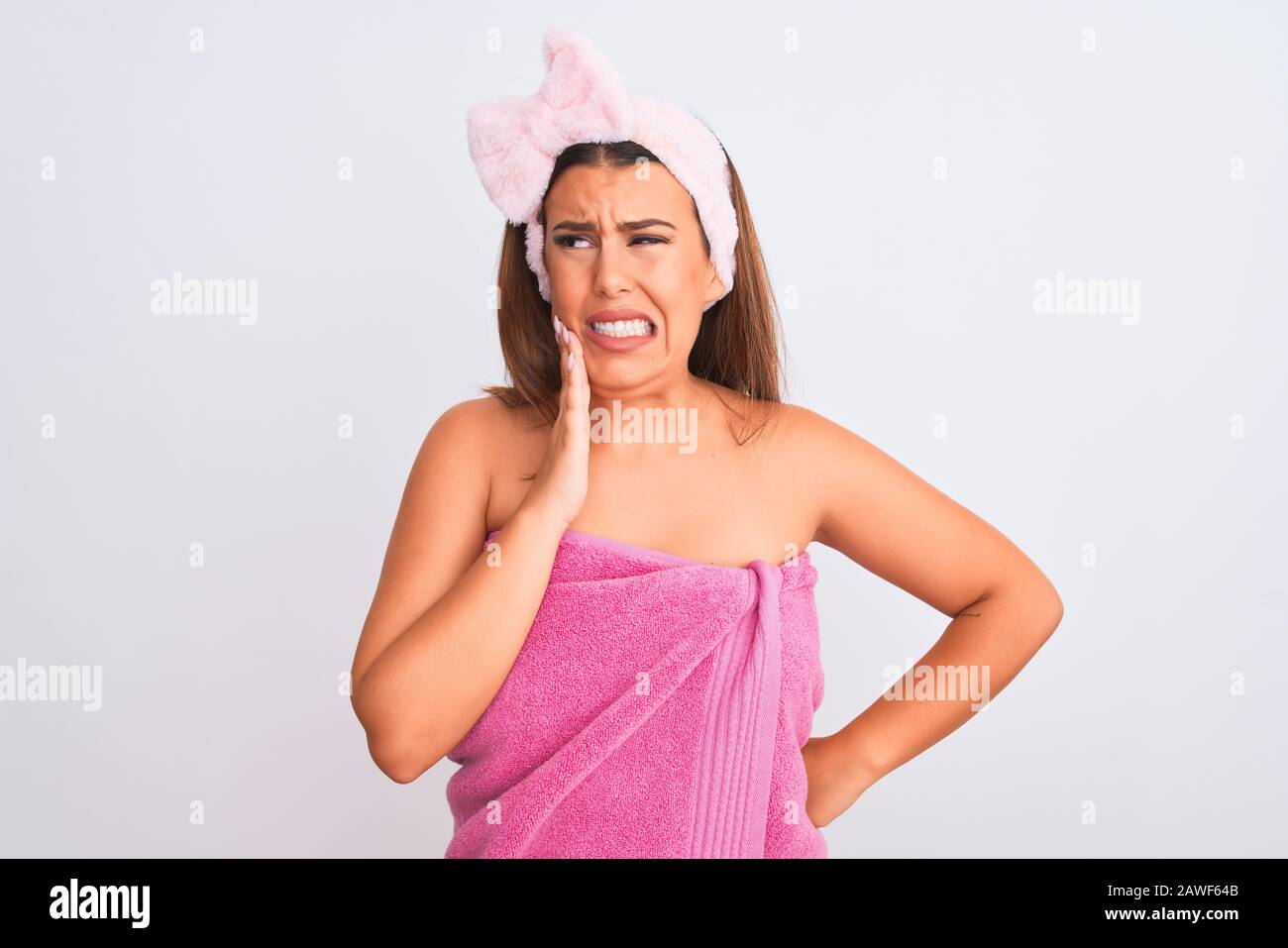 Beautiful young woman wearing pink shower towel and beauty headband over white background ...