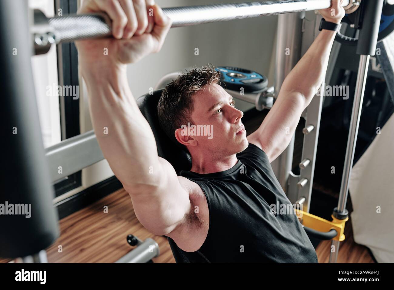 Determined young bodybuilder lifting barbell when sitting on gym bench ...