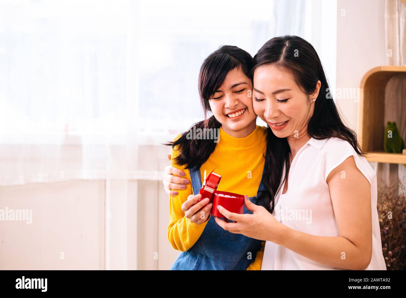 Smiling happy Asian teenage daughter and Asian middle-aged mother hugging together in indoor ...