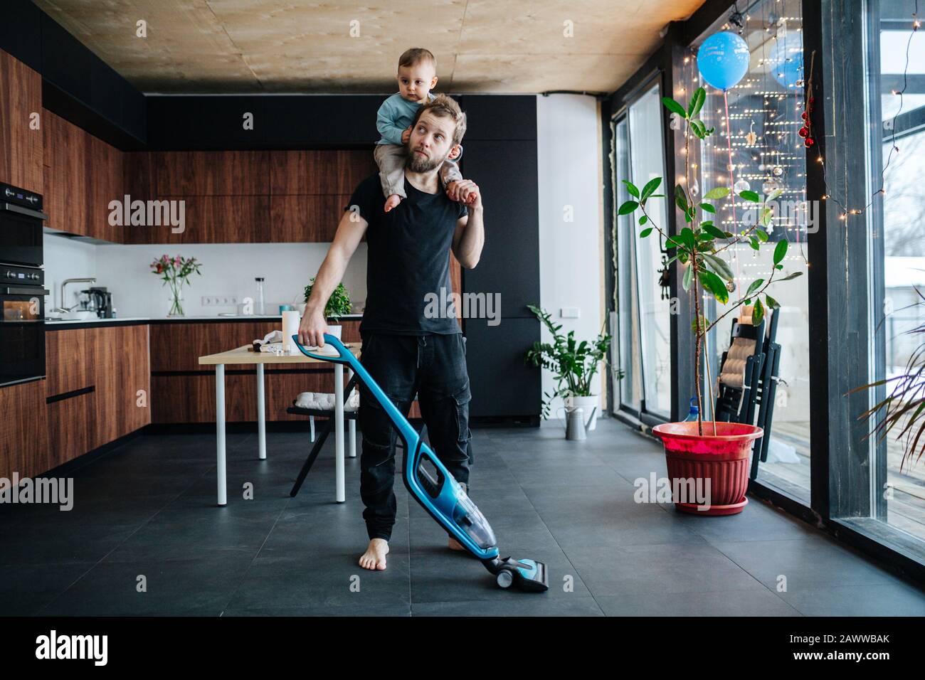 Young father vacuums apartment floor with his baby riding on his neck ...