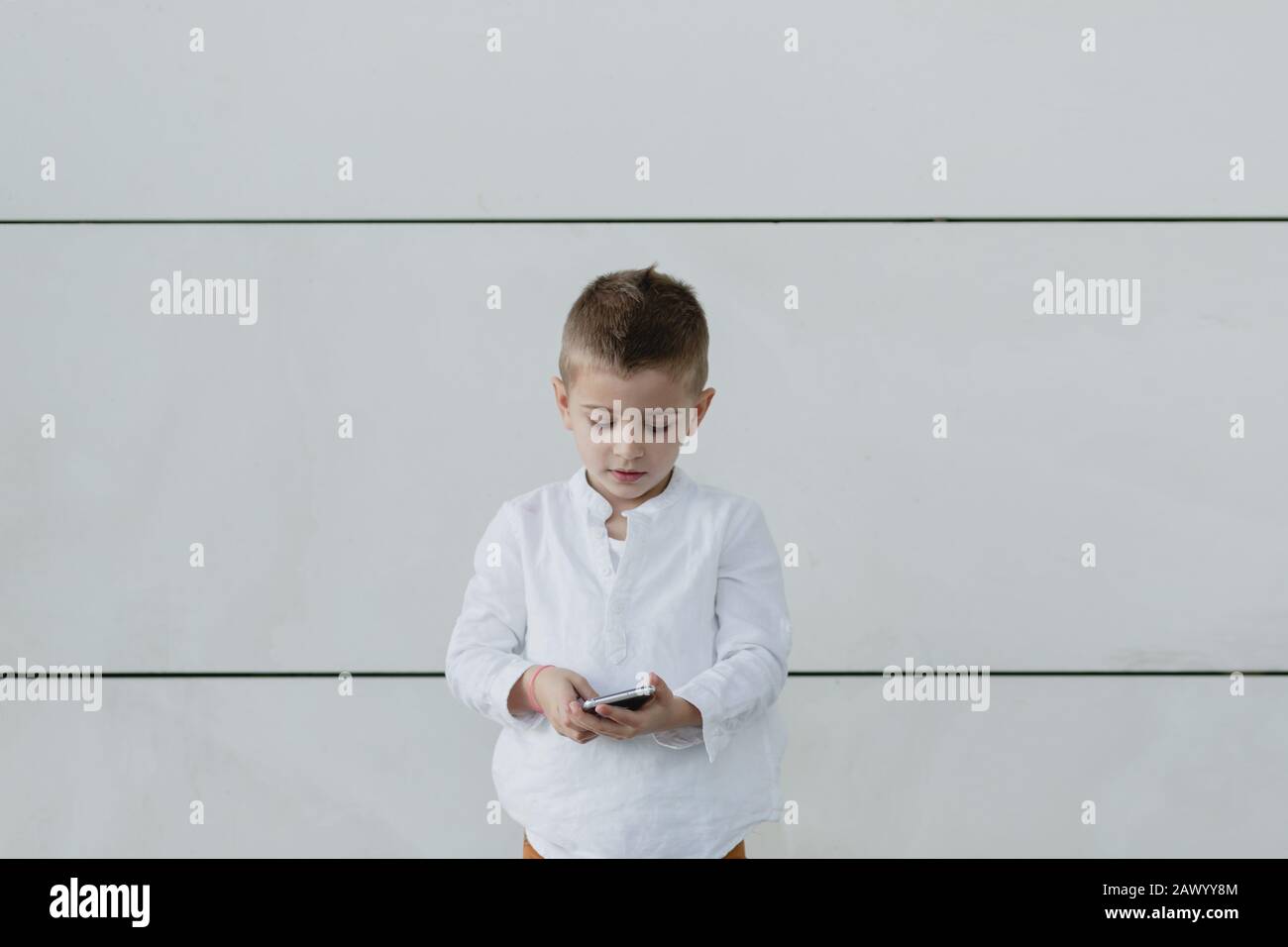 Boy looking down, playing on his phone with a white wall in the ...