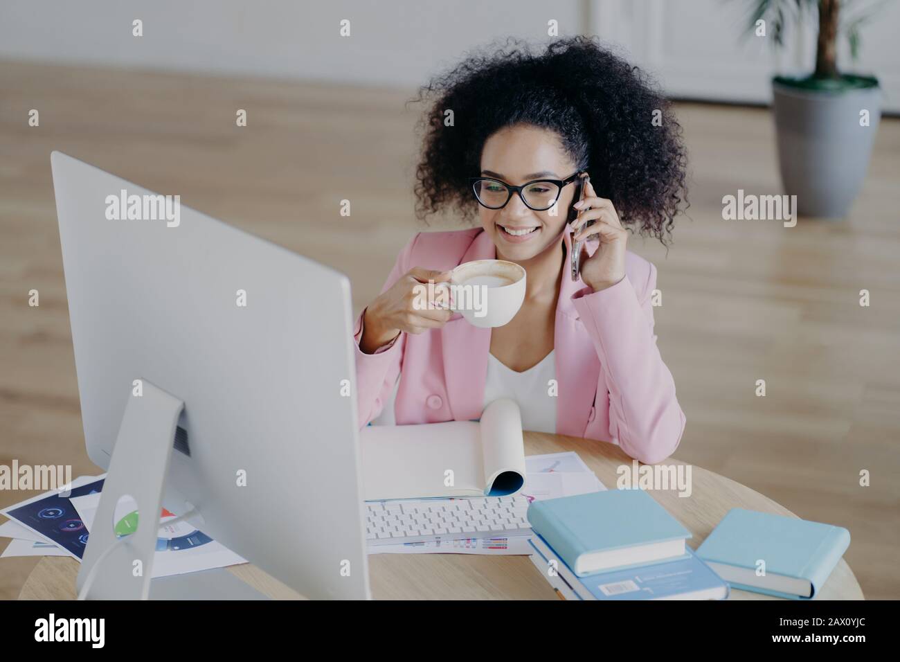 Glad dark skinned business lady looks happily at computer, drinks fresh ...