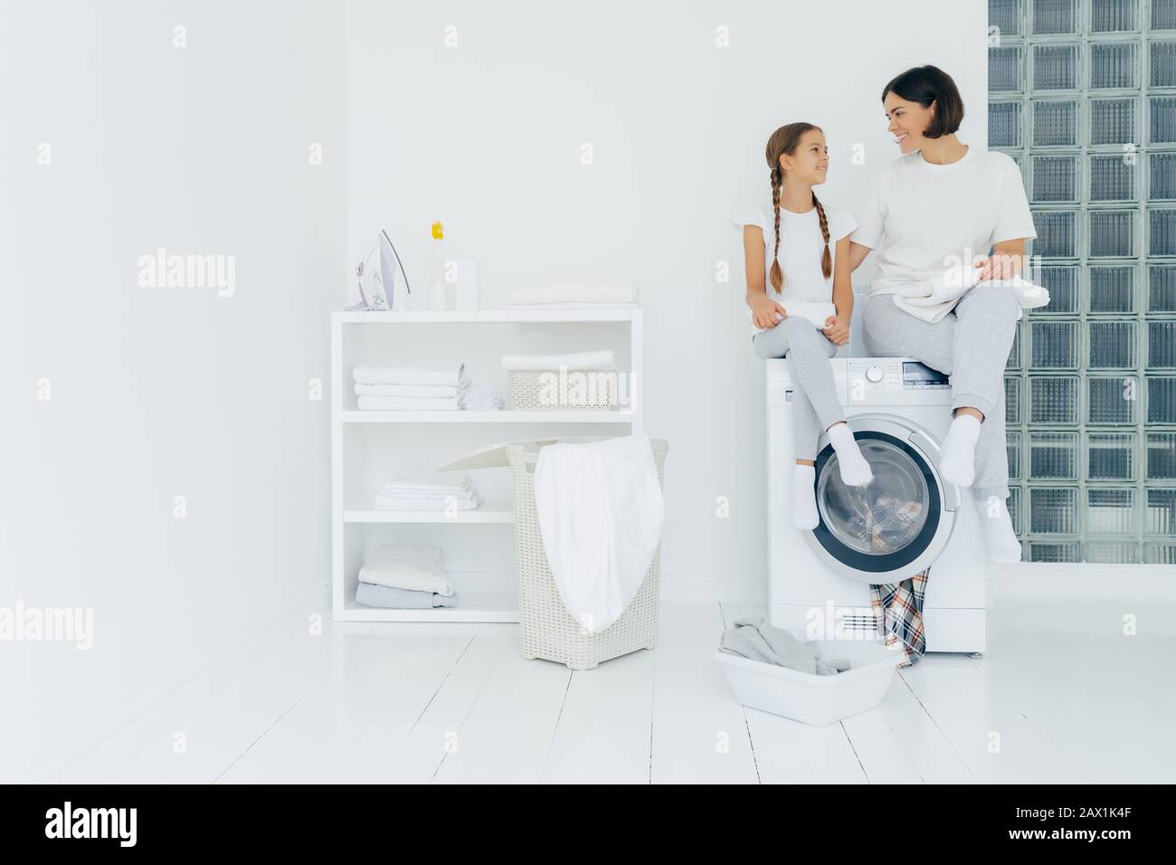 Housewife and small child sit on top of washing machine, holds freshly ...