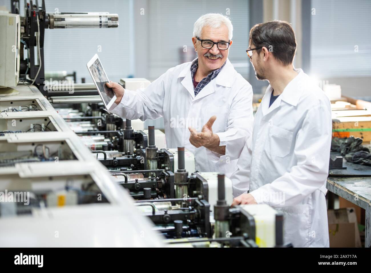 Positive senior inspector in lab coat talking to colleague while using ...