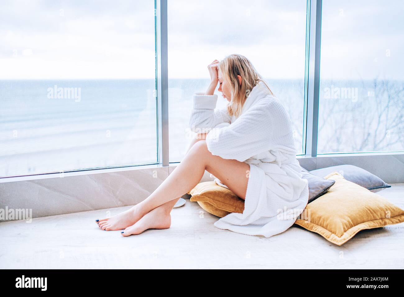 Woman in white bathrobe sitting on a floor near large window holding ...