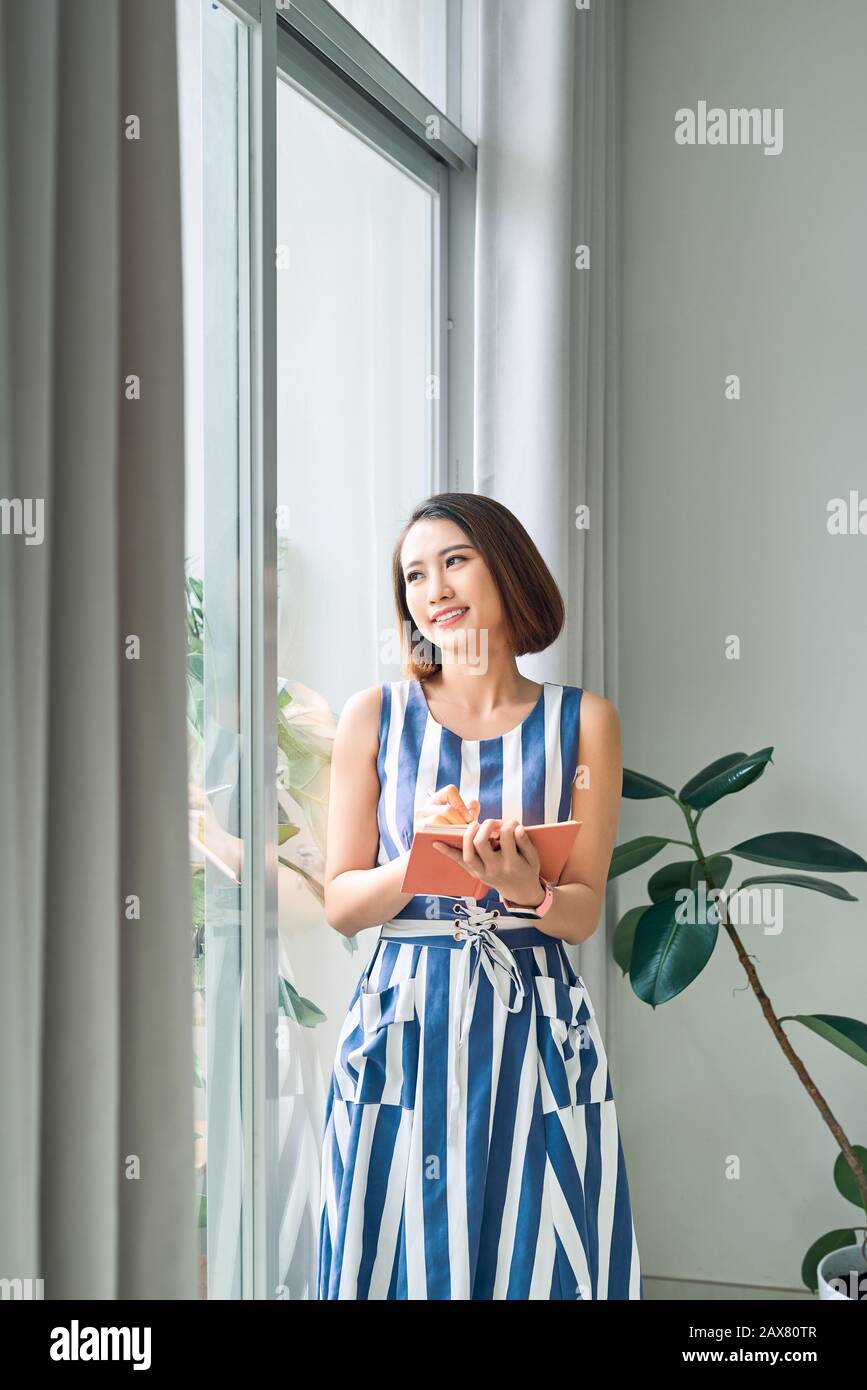 Portrait of young Asian woman standing, writing in notebook, looking ...