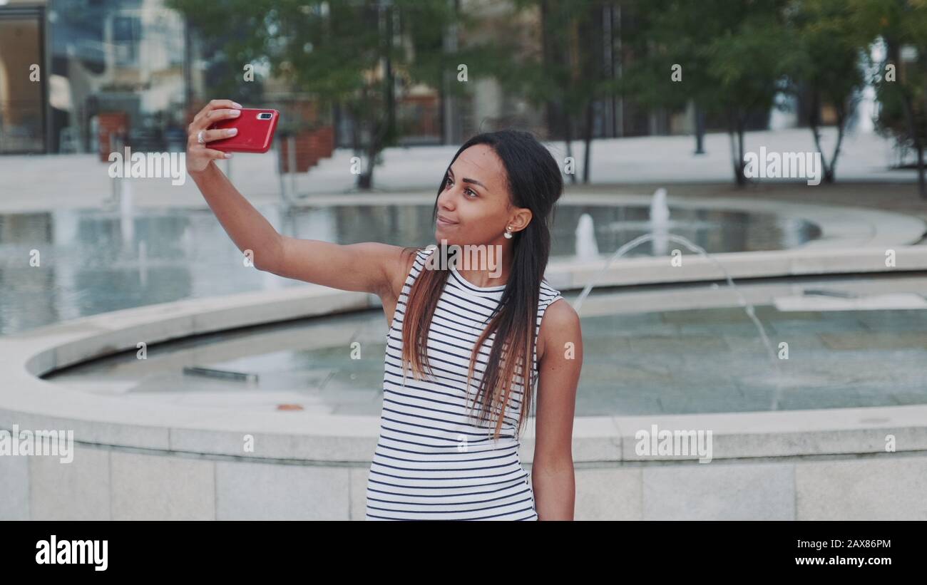 Cheerful african girl taking selfie in front of fountains in city ...