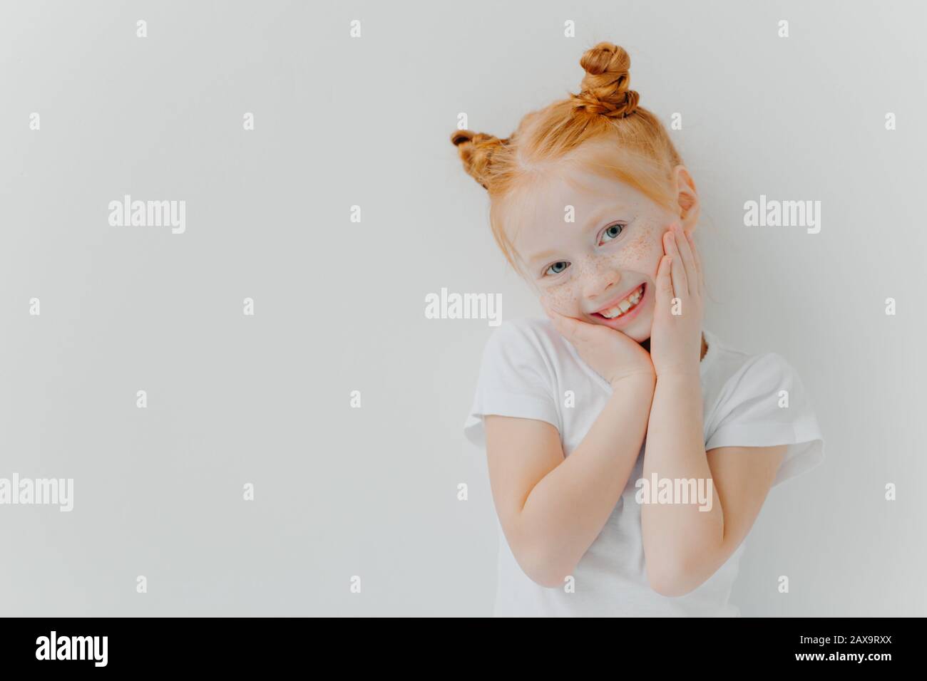 Happy small redhead girl tilts head, keeps hands on cheeks, smiles ...