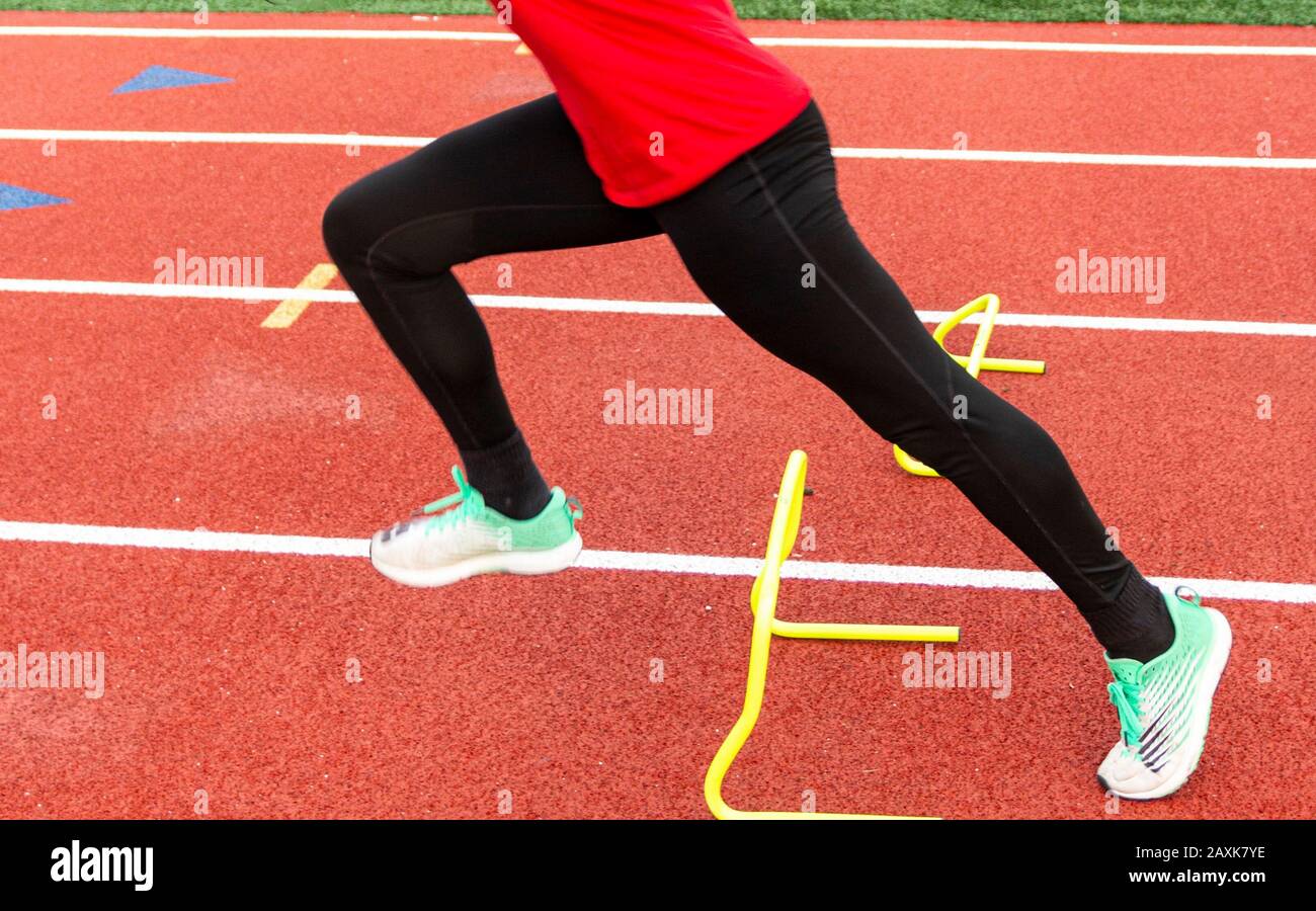 The legs of a high school runner performing a sprinters running drill over yellow mini hurdles ...