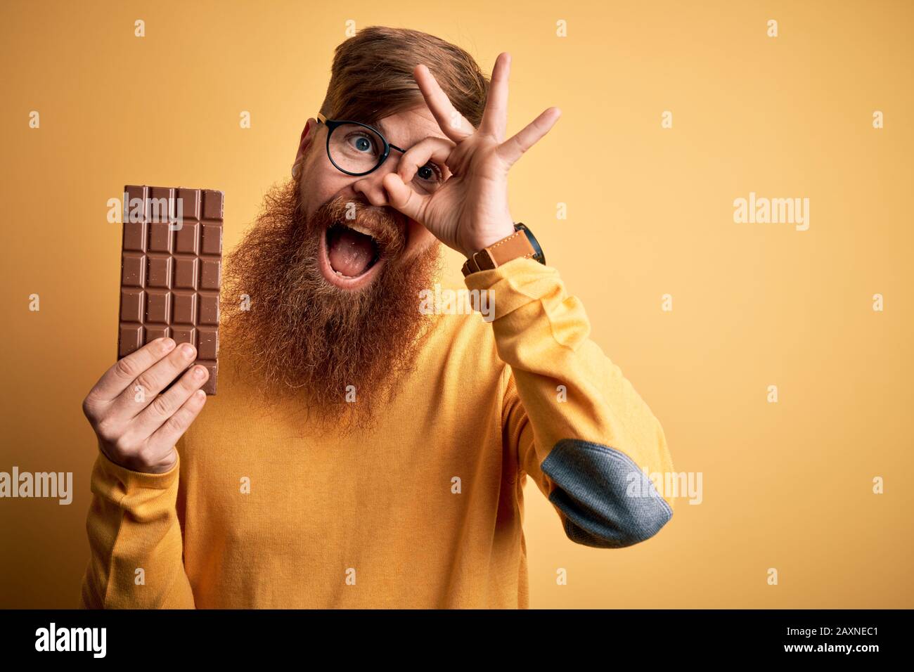 Irish redhead man with beard eating a sweet chocolate bar over yellow ...