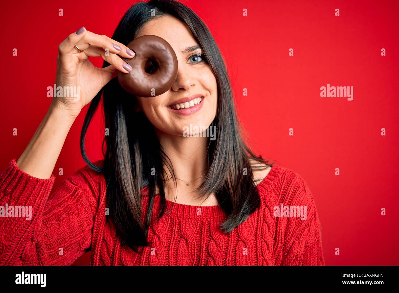 Young brunette woman with blue eyes eating chocolate doughnut over red ...