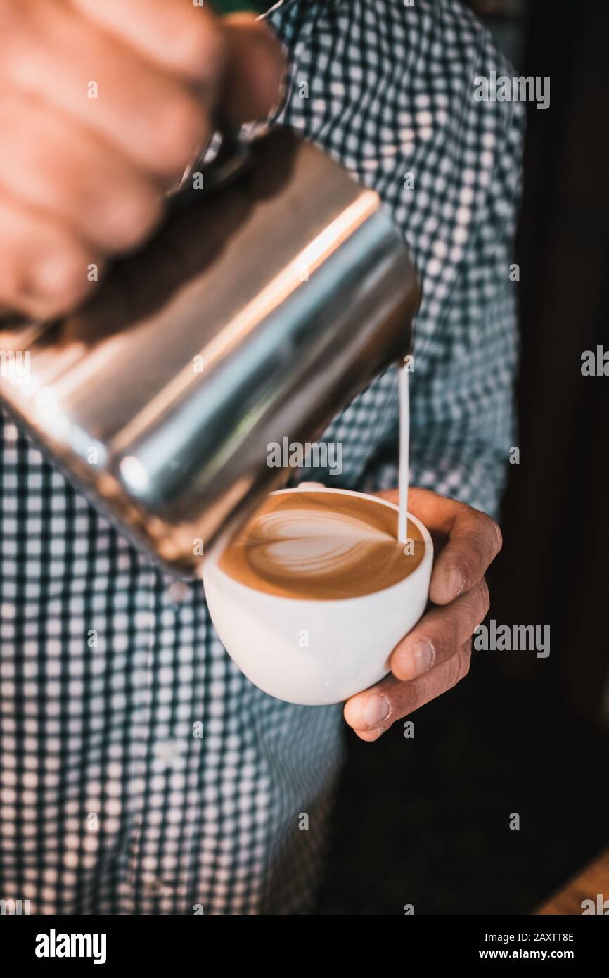 Vertical shot of a man pouring a foamed milk into a cup of cappuccino ...