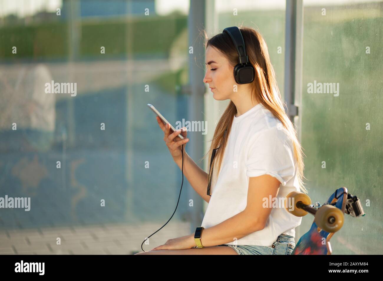 Stylish young girl sitting on bus stop with her longboard, listening ...