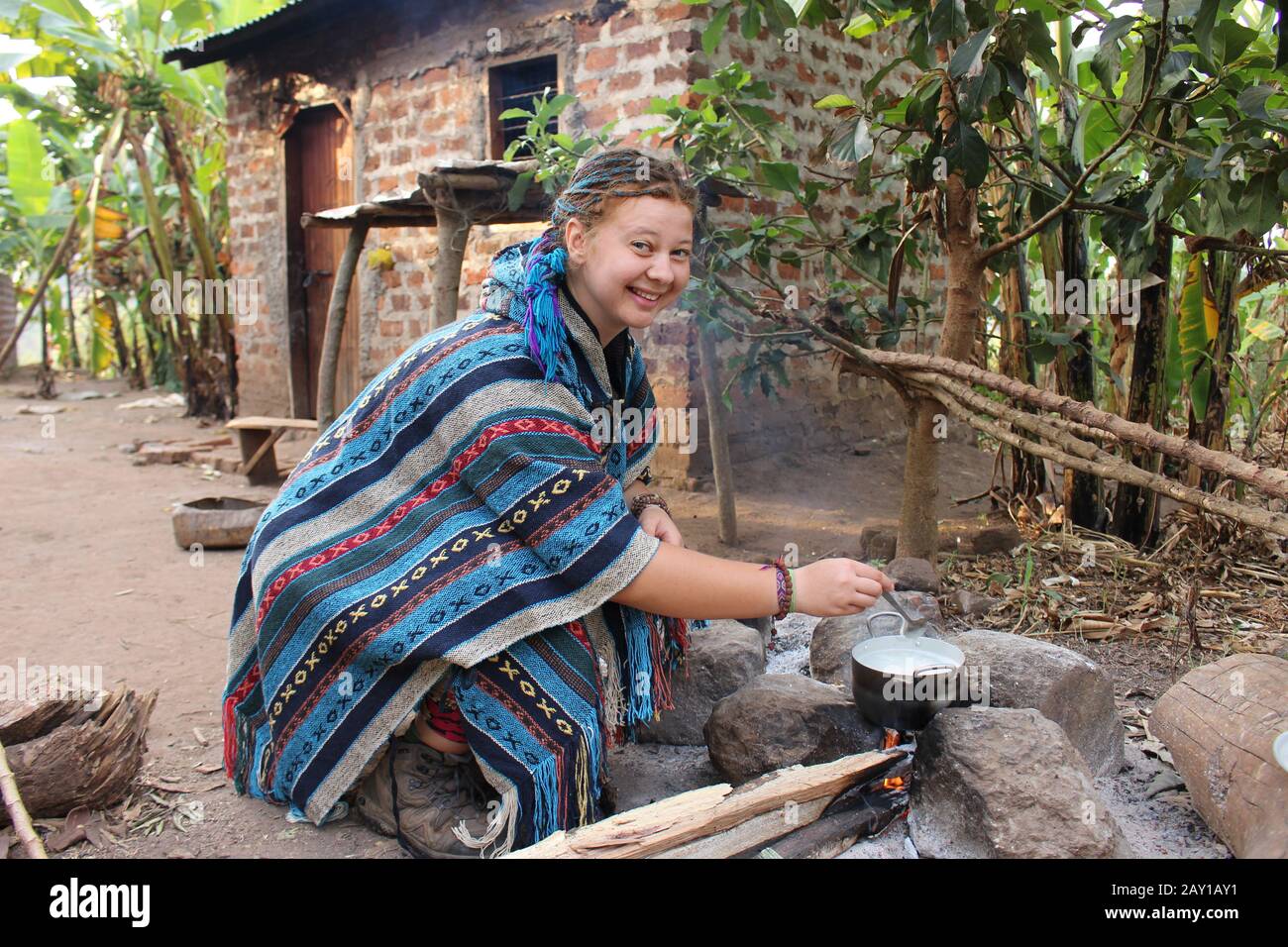 Young hippie woman in a blue poncho cooking on a campfire campground in ...