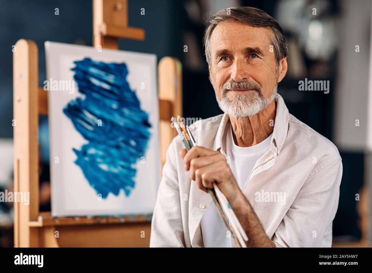 Portrait of bearded painter sitting against his picture and looking ...