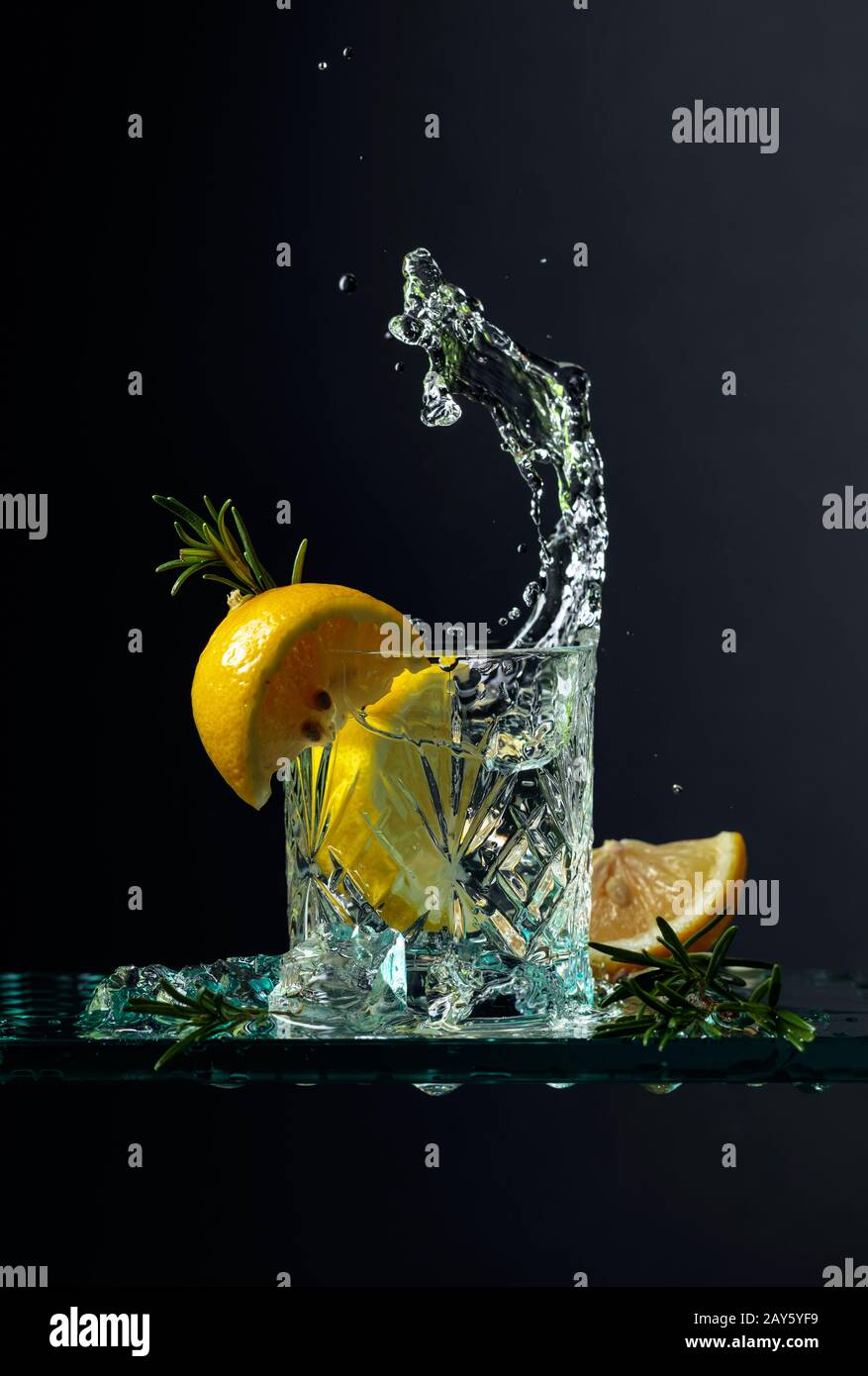 Cocktail gin-tonic with lemon and rosemary. Carbonated drink with ice pieces on a glass table ...