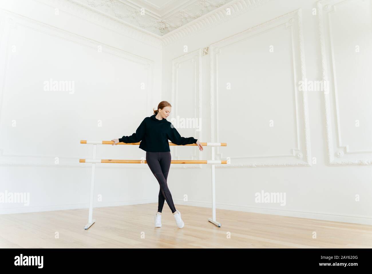 Classic ballet dancer has stretching workout, stands near ballet barres, wears black sweatshirt ...