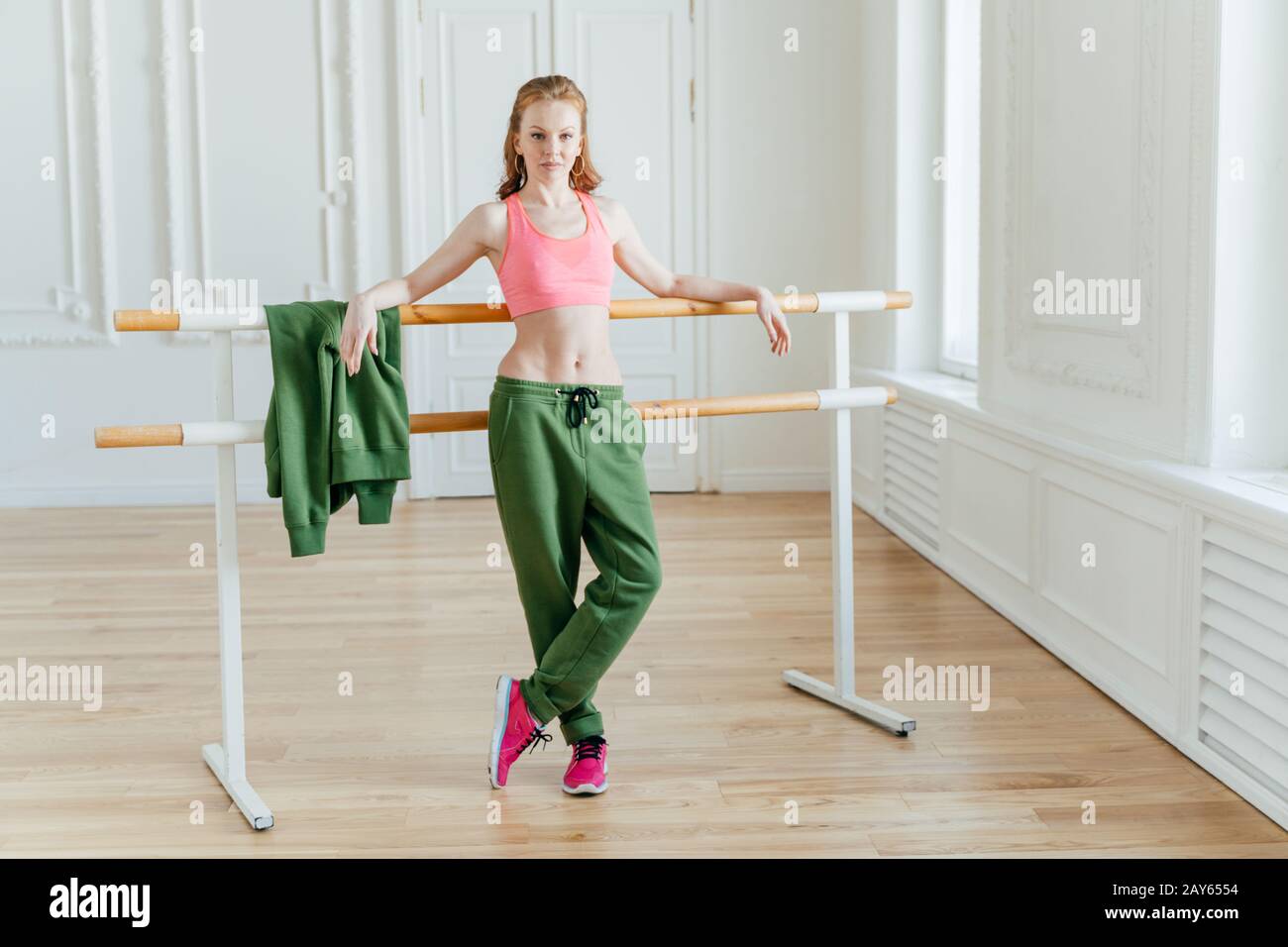 Slim beautiful female ballet dancer leans at ballet barre, wears pink ...
