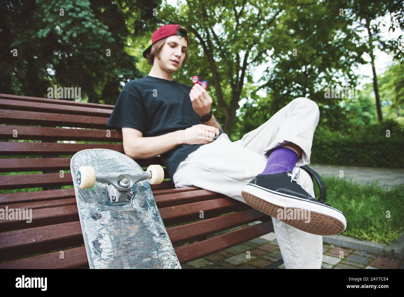 Long-haired hipster skater in a cap sits on a bench and spins on a spin ...