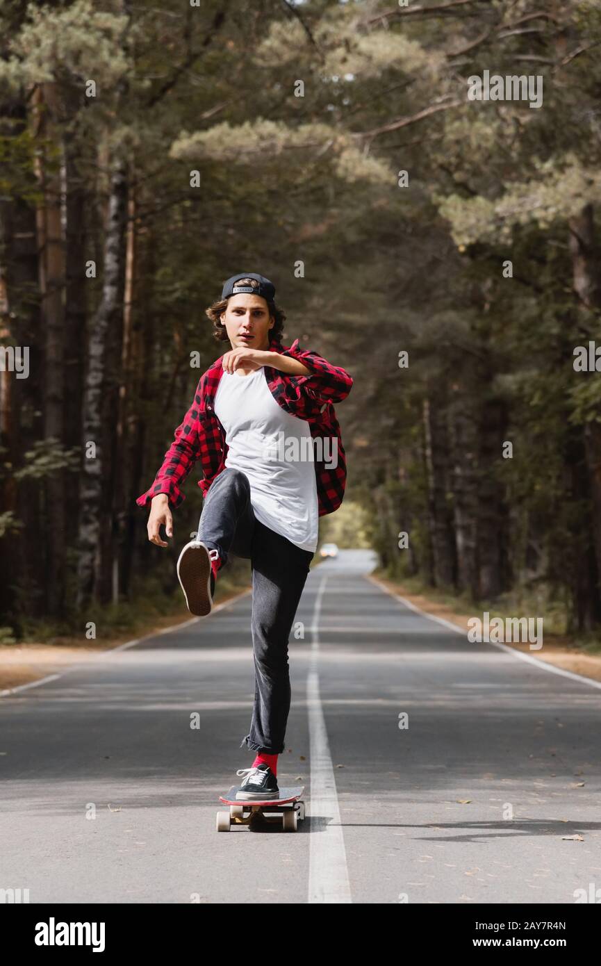 A young hipster in a cap and plaid shirt is riding his longboard on a ...